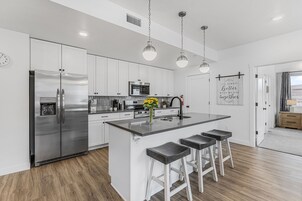 Uma cozinha moderna com armários brancos, eletrodomésticos de aço inoxidável, uma ilha central com banquetas e iluminação pendente.