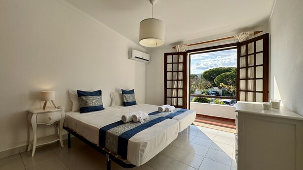 Quarto com cama, mesa de cabeceira, luminária, ar condicionado e vista pela grande janela.