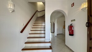 Uma escadaria com degraus de madeira e paredes brancas, conduzindo a uma porta em arco.