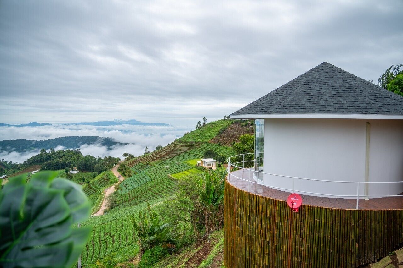Um edifício moderno com fachada branca e telhado escuro, rodeado por campos verdes em socalcos e um céu nublado.