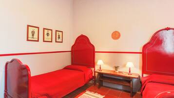 Quarto com duas camas vermelhas, uma mesinha de cabeceira de madeira com duas luminárias e três quadros na parede.