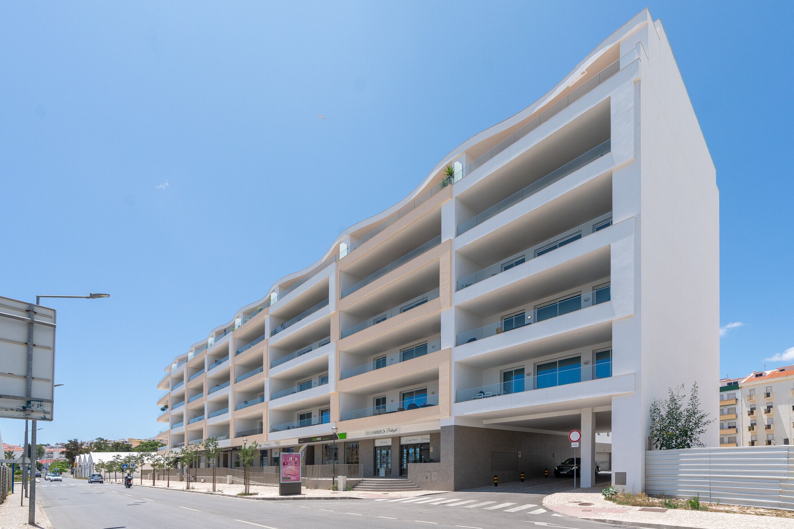 Um hotel com vários andares e design moderno, com varandas em cada andar, área de estacionamento coberta e um céu azul claro ao fundo.