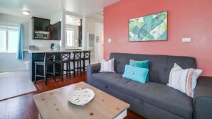 Sala de estar com sofá cinzento, mesa de centro e cozinha ao fundo.