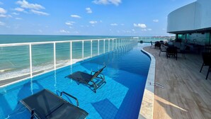 Uma área na piscina com vista para o oceano, um guarda-corpo branco e um edifício moderno com fachada revestida de azulejos.