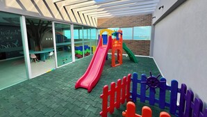 Espaço infantil com escorregador e estruturas de escalada, parede de vidro com um mural de árvore, e vista para o mar através de amplas janelas.
