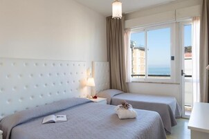 Quarto de hotel com uma cama grande, uma cama menor, uma mesa de cabeceira com abajur e vista para o mar através de uma janela ampla.