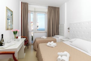 Quarto de hotel com duas camas, uma escrivaninha com cadeira, uma mesinha com uma garrafa e copos, e vista para o mar pela janela.