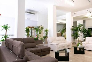 Uma sala de estar moderna com um sofá modular, uma poltrona branca e uma mesa de centro de vidro. Há várias plantas em vasos e uma escada ao fundo.