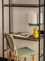 Uma cadeira de vime com assento e encosto trançados, uma mesa de madeira com um abajur amarelo e um livro aberto sobre a mesa.