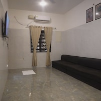 Quarto com televisão de tela plana fixada na parede, ventilador de teto, janela com cortinas, sofá de cor escura e piso de azulejos.
