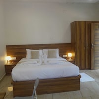 Cama bem arrumada com lençóis brancos e duas almofadas, com luminárias de cabeceira de cada lado, num quarto com piso de madeira e um guarda-roupa grande.