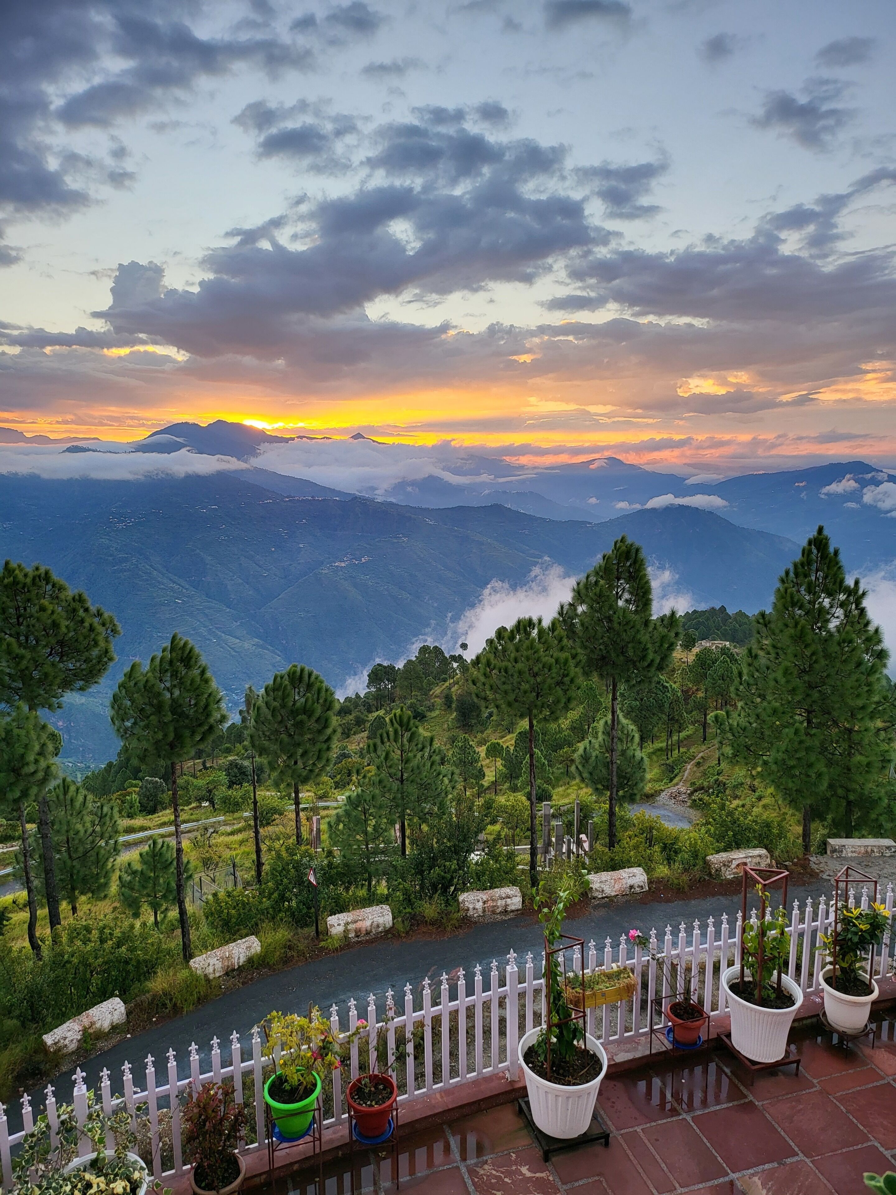 Terraço com plantas em vasos, cerca branca de piquete e vista para uma paisagem montanhosa ao pôr do sol.