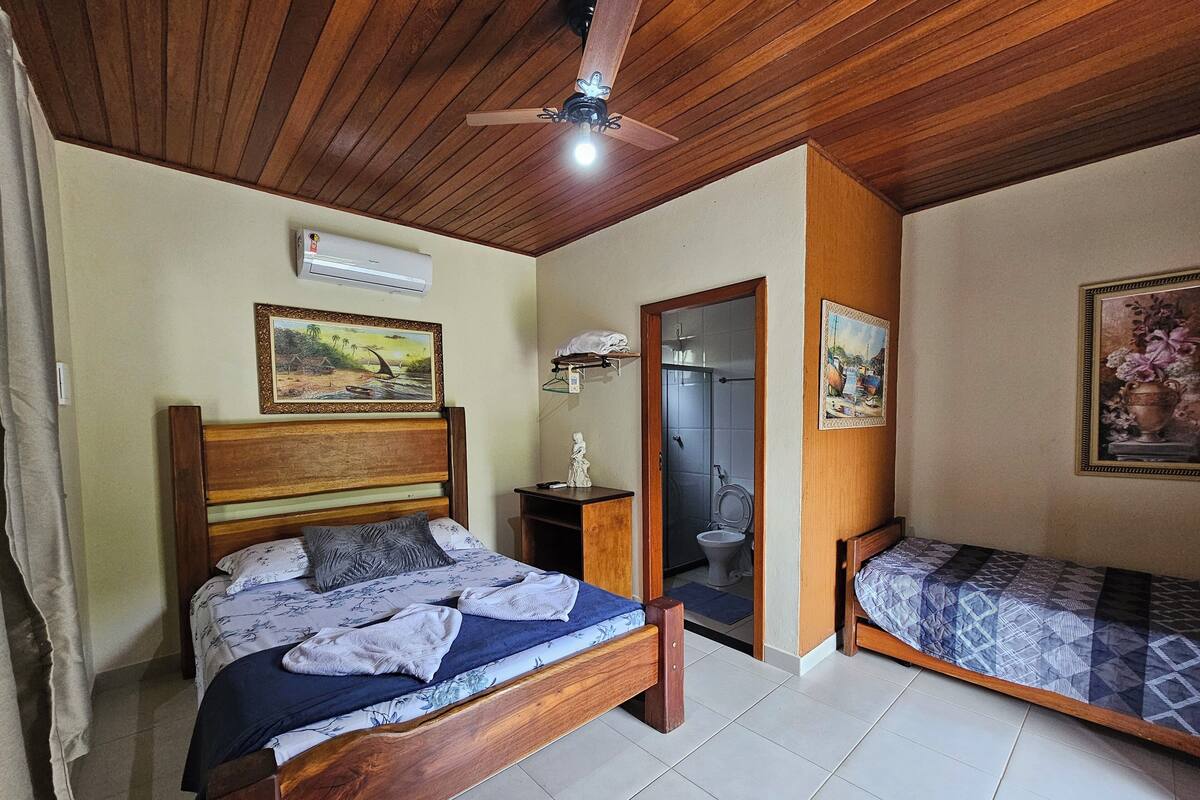 Quarto com teto de madeira, cama com colcha azul e branca estampada, mesa de cabeceira com abajur, uma pequena estátua e um quadro na parede.