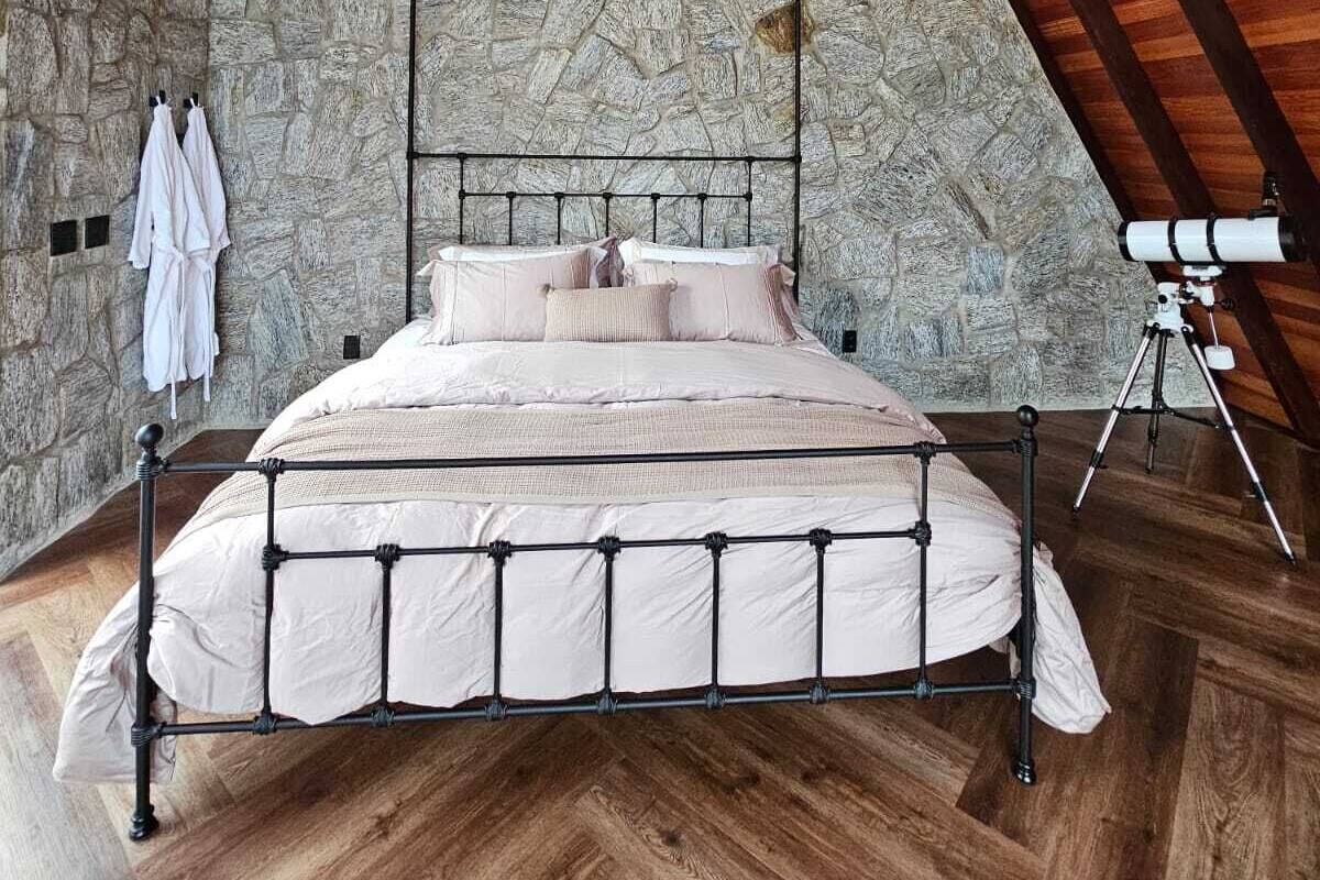 Quarto com parede de pedra, teto de madeira, cama com estrutura de metal e um telescópio sobre tripé.