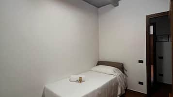 Quarto pequeno com uma cama de solteiro, teto de madeira, paredes brancas e chão ladrilhado. Um aparelho de ar condicionado está fixado na parede acima da cama.