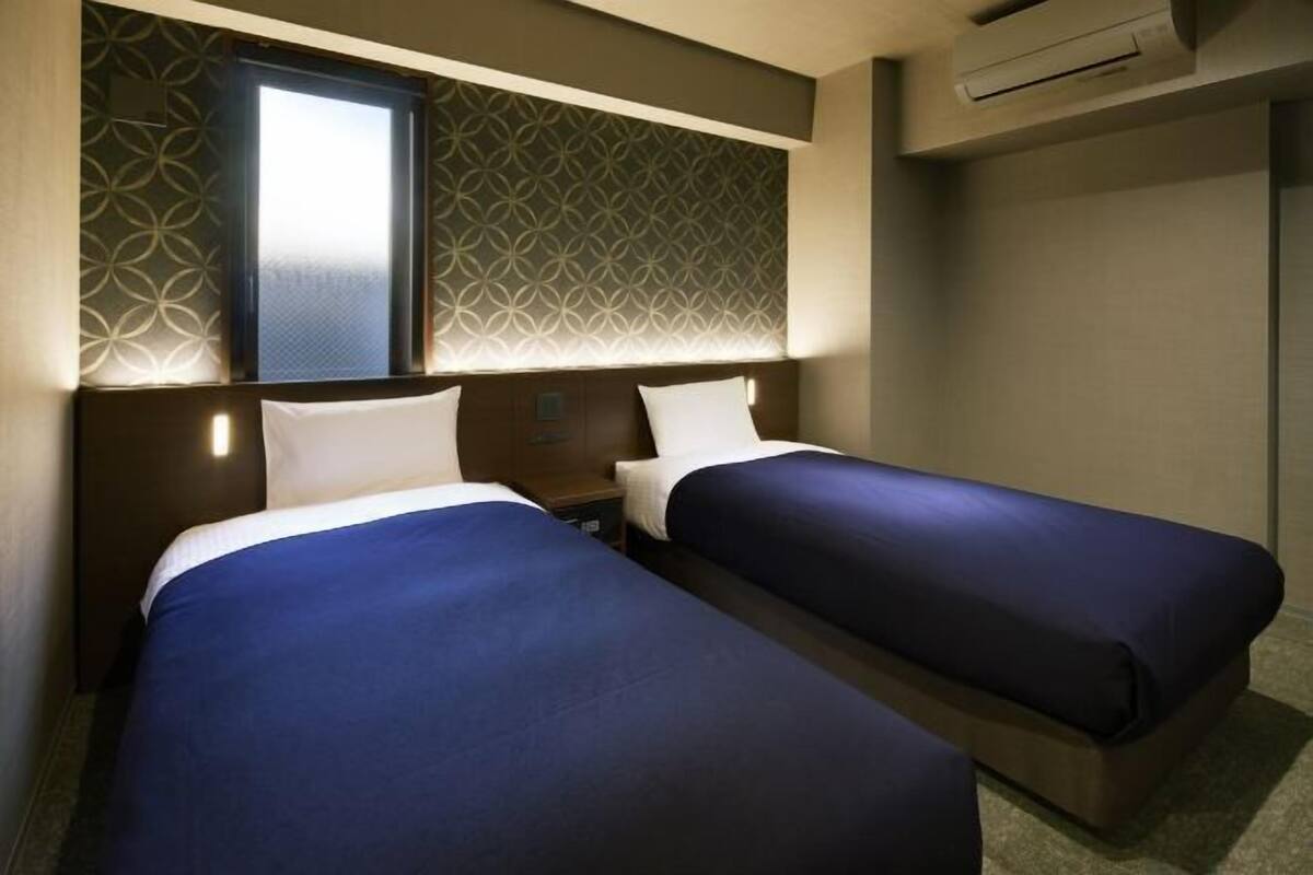 Quarto de hotel com duas camas, mesa de cabeceira, ar condicionado fixado na parede e papel de parede estampado.