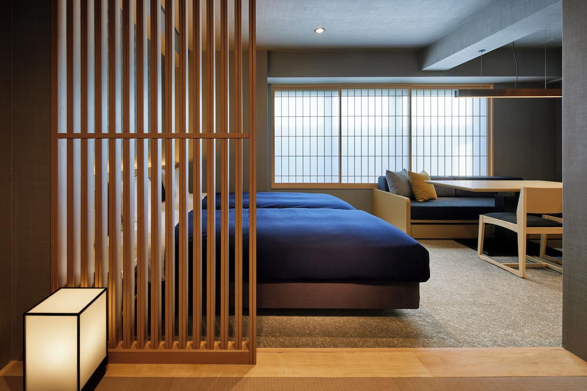 Quarto de hotel moderno com uma janela ampla, uma cama, uma mesa de trabalho e um painel com ripas verticais.