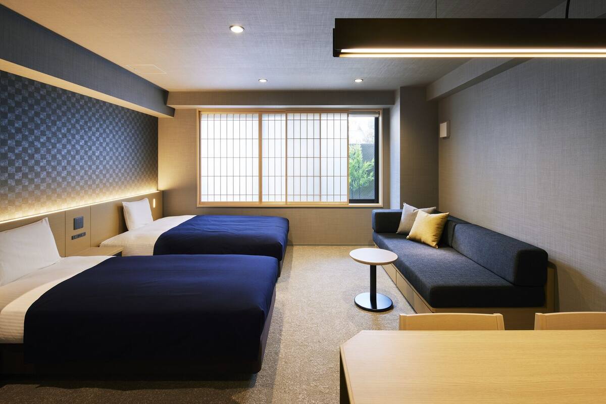 Quarto de hotel com duas camas, um sofá, uma mesa redonda pequena e uma janela grande que oferece luz natural.