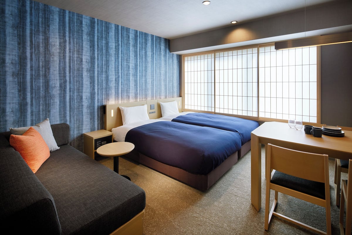 Quarto de hotel com cama, sofá, mesa pequena e janela com persianas.