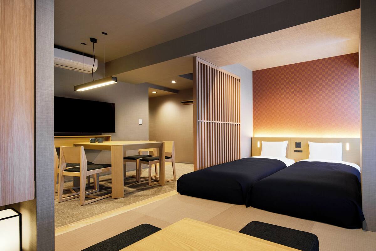 Quarto de hotel moderno com cama, escrivaninha, cadeiras e televisão.