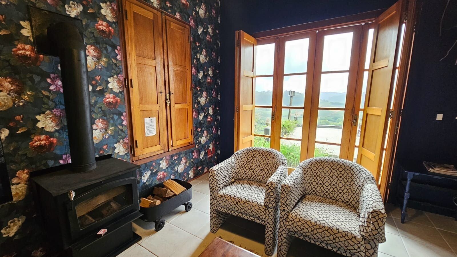 Um quarto aconchegante com lareira, papel de parede floral, duas poltronas estampadas e uma janela grande com vista.