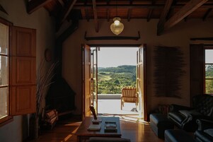 Uma sala de estar acolhedora com teto de madeira, lareira, vista para o campo e um espaço de estar com sofá e poltrona.
