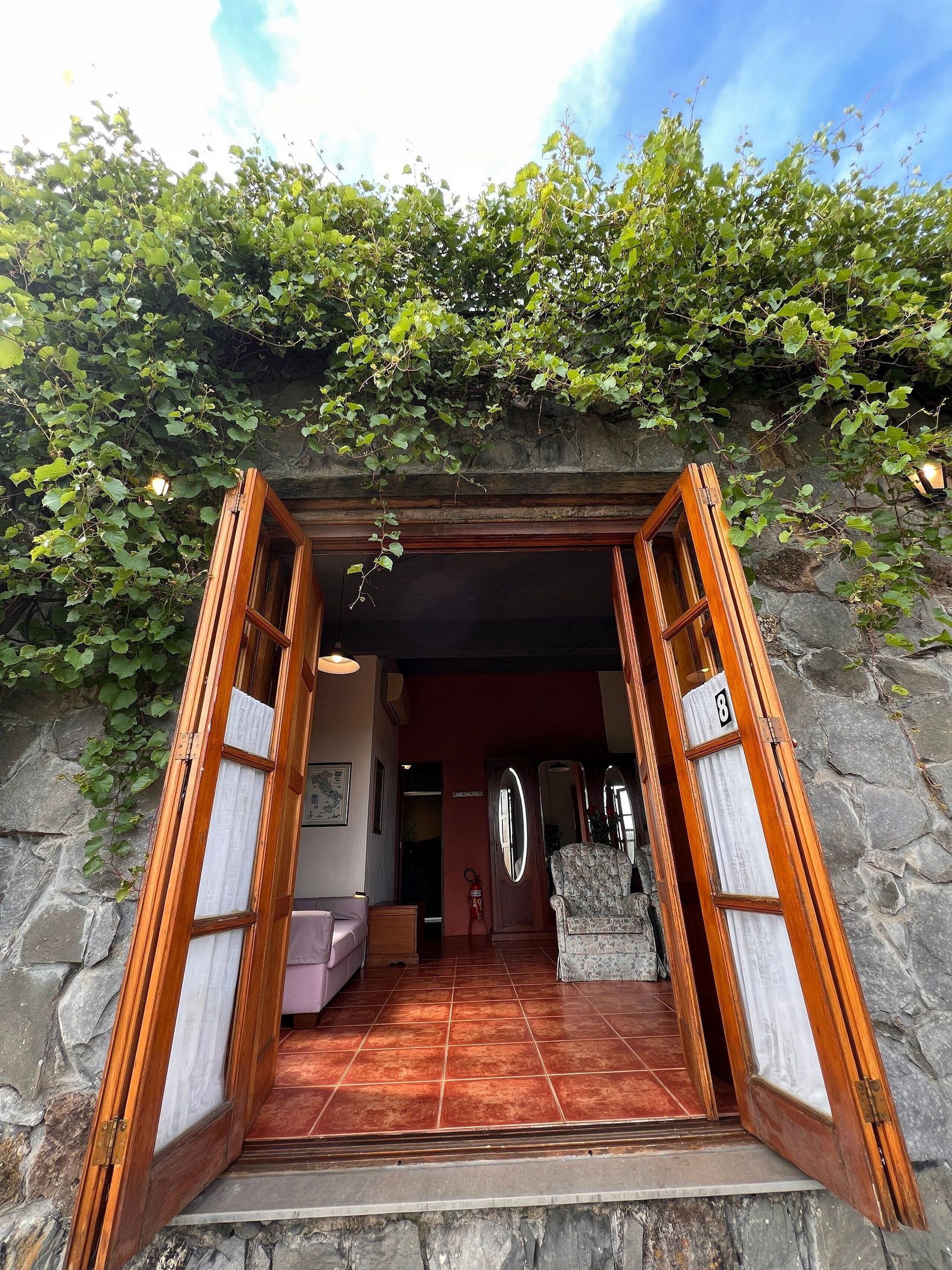 Entrada com paredes de pedra, contraportas de madeira, soalho em mosaico e vista para um quarto com cama e poltrona.