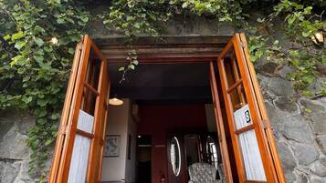 Entrada com paredes de pedra, contraportas de madeira, soalho em mosaico e vista para um quarto com cama e poltrona.