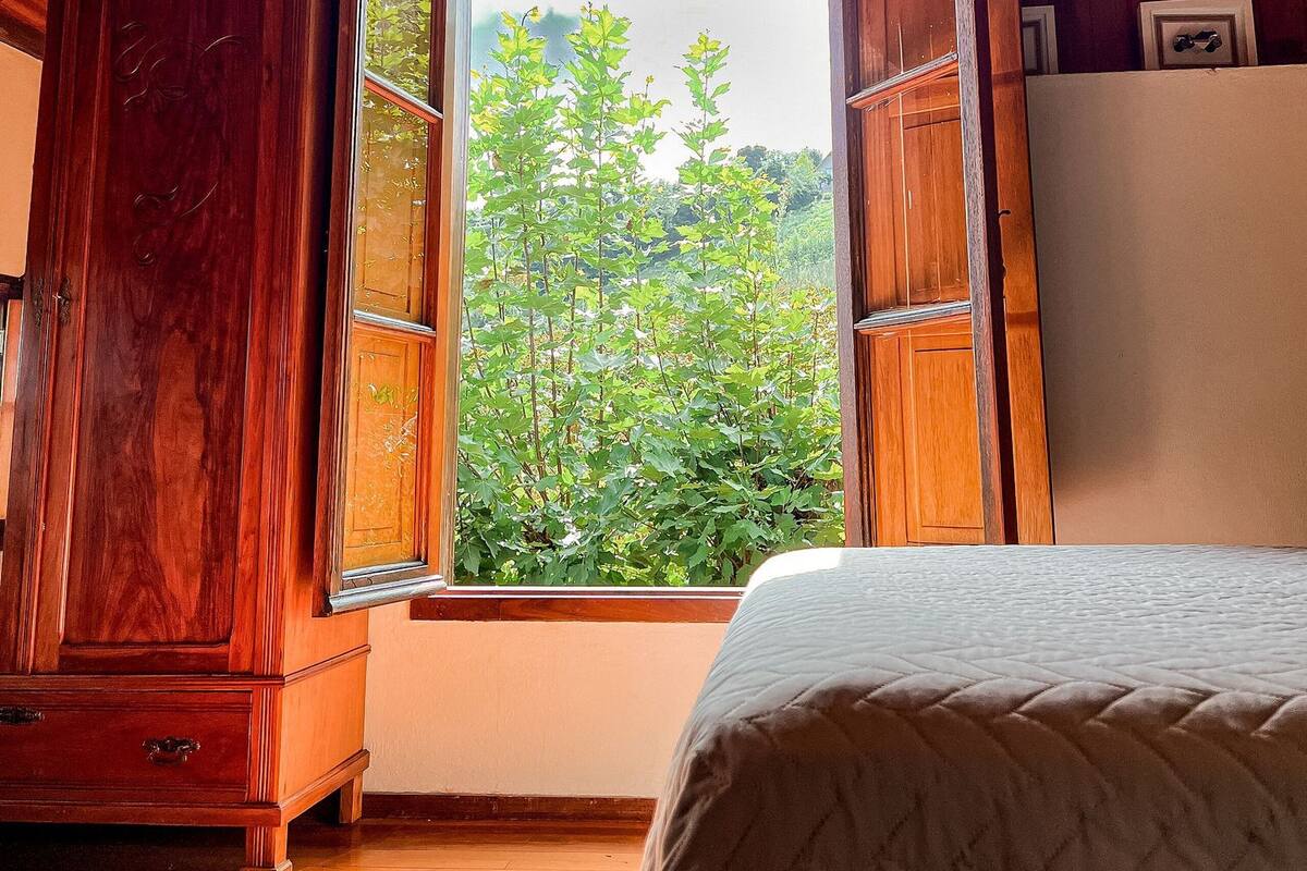 Quarto com teto de madeira, uma janela ampla com vista para o verde, uma cama e um guarda-roupa.