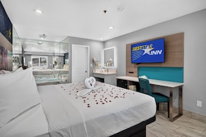 Quarto de hotel com cama, escrivaninha, televisão e banheiro visível através de um painel de vidro.