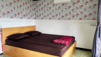 Uma cama de solteiro com estrutura de madeira e colchão marrom, coberta por um cobertor vermelho, e papel de parede com padrão floral.