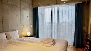 Quarto de hotel com uma cama grande, uma mesa de cabeceira com abajur, vista de um edifício pela janela e uma cadeira perto da janela.