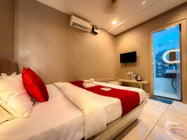 Quarto de hotel com cama, almofada vermelha, colcha branca, manta vermelha e branca, televisão fixada na parede, banheiro com espelho e chuveiro.