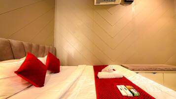 Quarto de hotel com cama, parede decorada em vermelho, cabeceira branca, travesseiro vermelho, toalha branca e uma mesinha com uma garrafa e um livro.