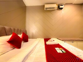 Quarto de hotel com cama, parede decorada em vermelho, cabeceira branca, travesseiro vermelho, toalha branca e uma mesinha com uma garrafa e um livro.