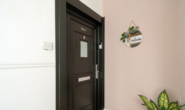 Uma porta marrom escura com placa de identificação '166A' e um letreiro de 'welcome' com uma planta na parede.