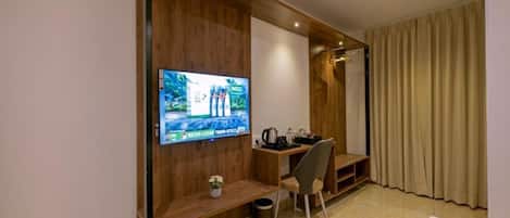 Quarto de hotel com televisão na parede, uma secretária com cadeira, uma planta pequena e uma janela com cortinas.