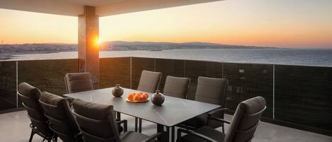 Uma área de jantar com mesa e cadeiras, uma tigela de frutas e vista para o pôr do sol sobre a água.