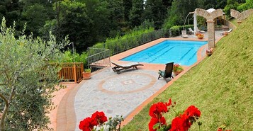 Piscina com deck de pedra, cercada por uma grade e vegetação.