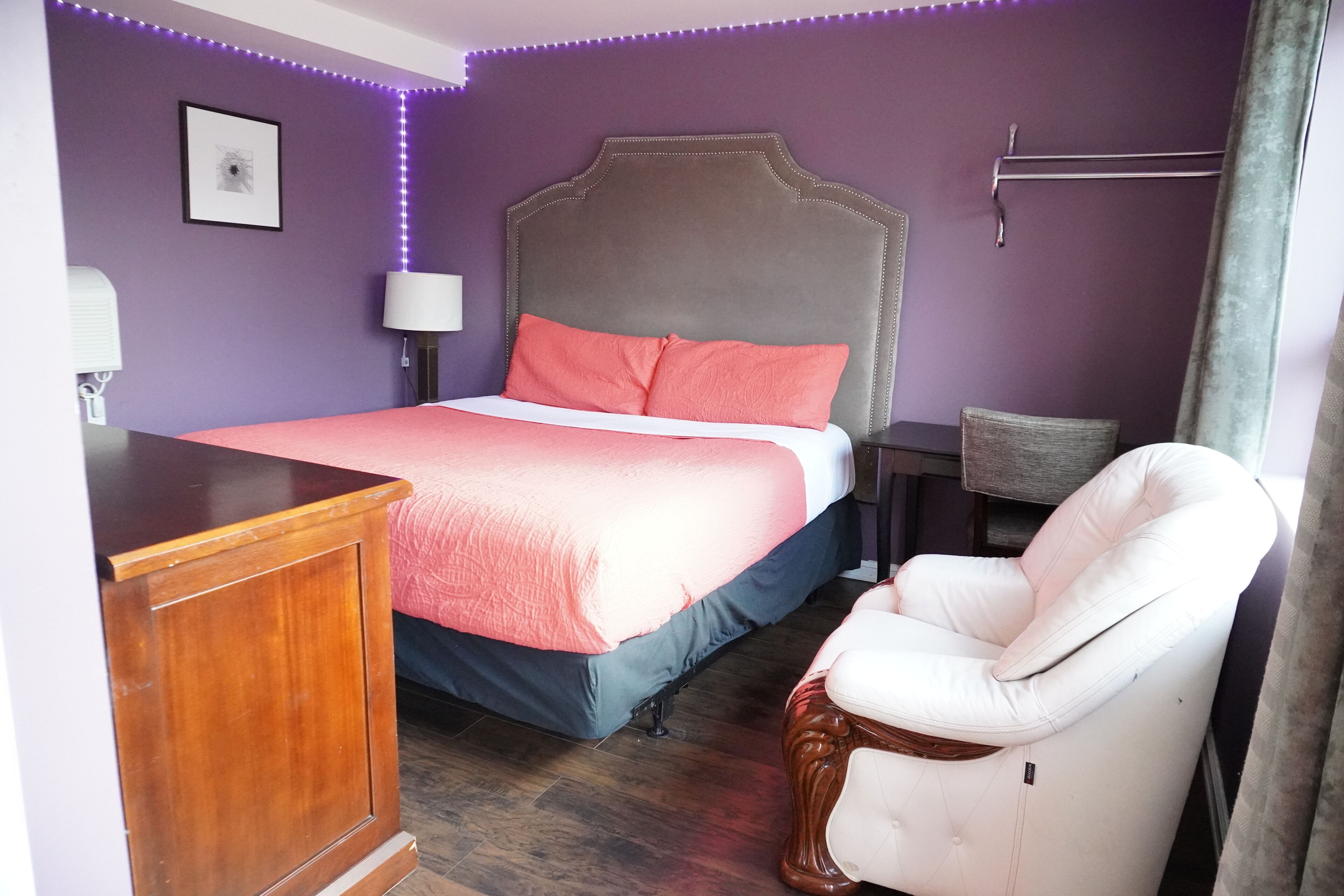 Quarto com cama, cómoda de madeira, poltrona branca e paredes roxas.