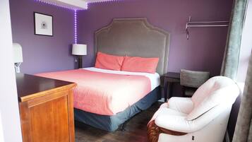 Quarto com cama, cómoda de madeira, poltrona branca e paredes roxas.
