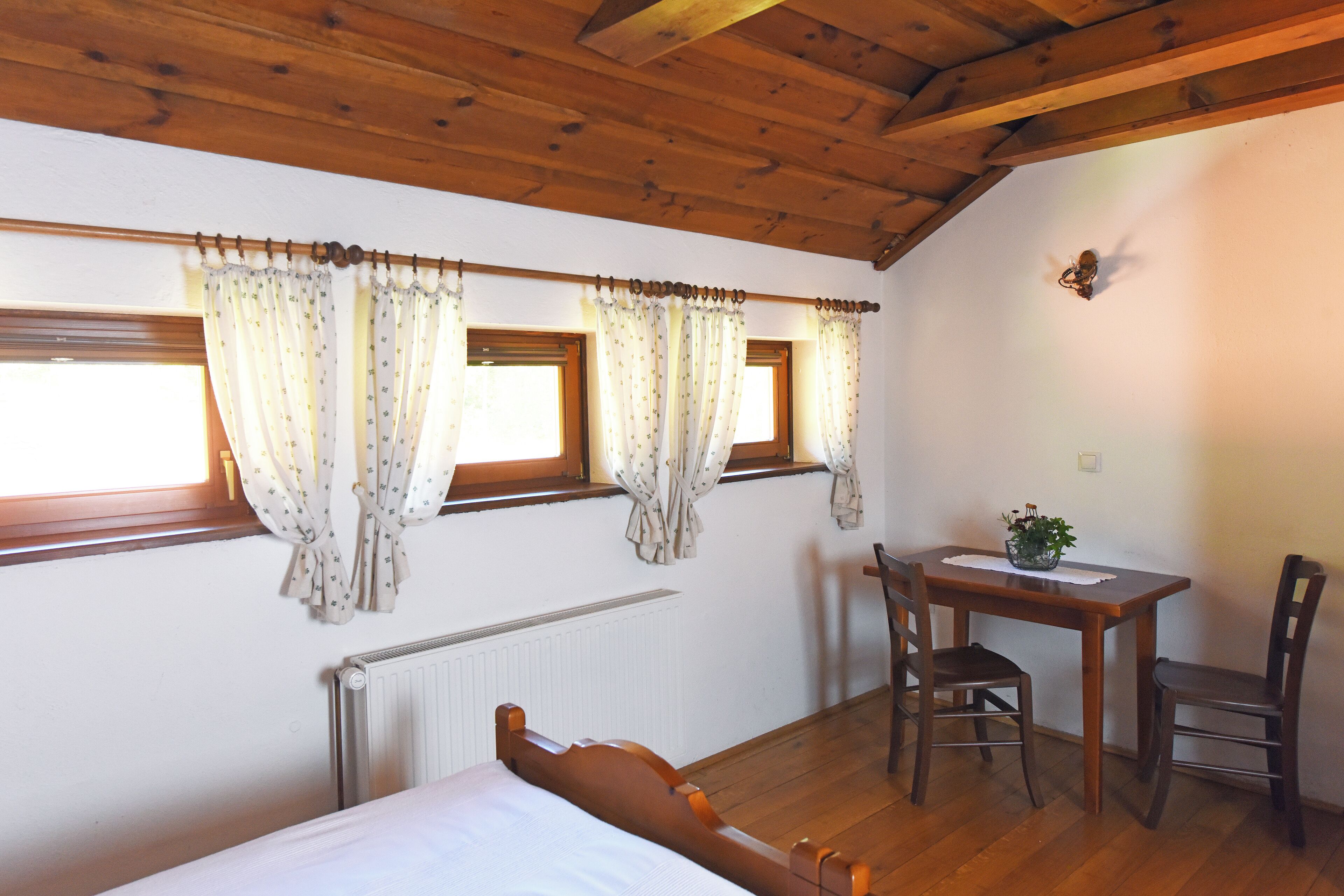 Quarto com cama de madeira, uma mesinha com cadeiras e uma janela com cortinas.