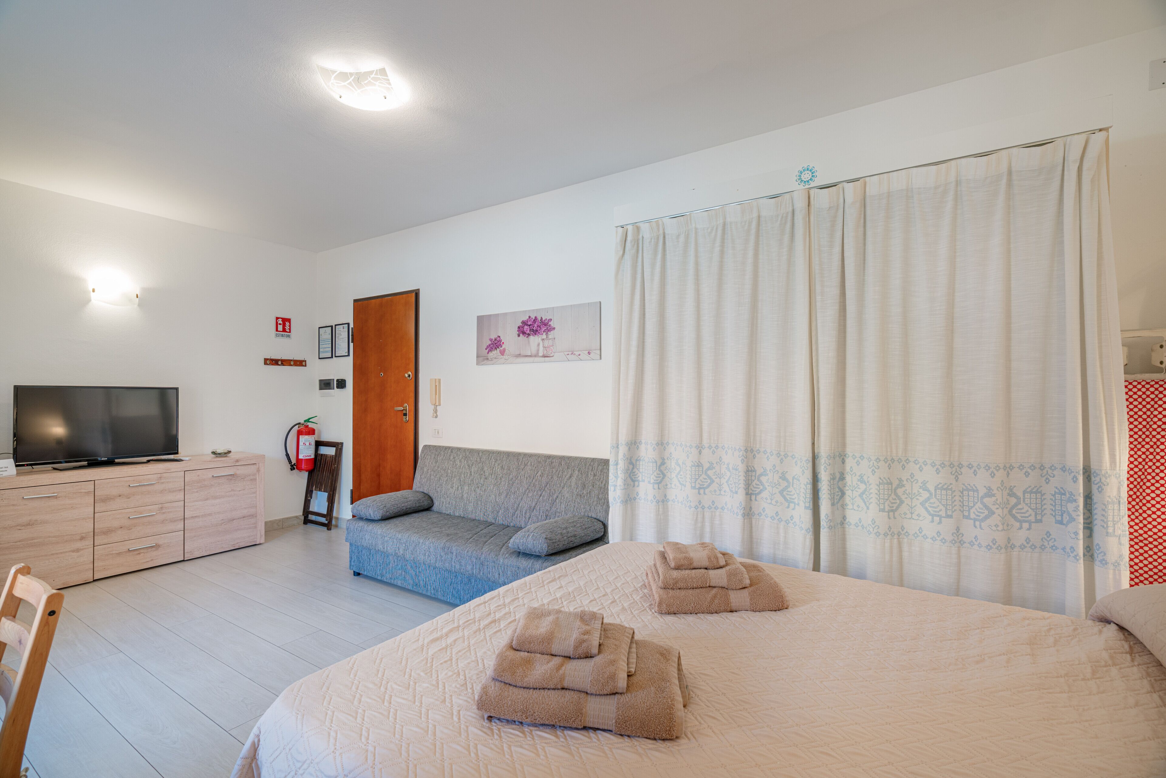 Quarto de hotel com uma cama, um sofá, uma televisão, um extintor de incêndio e uma cortina.