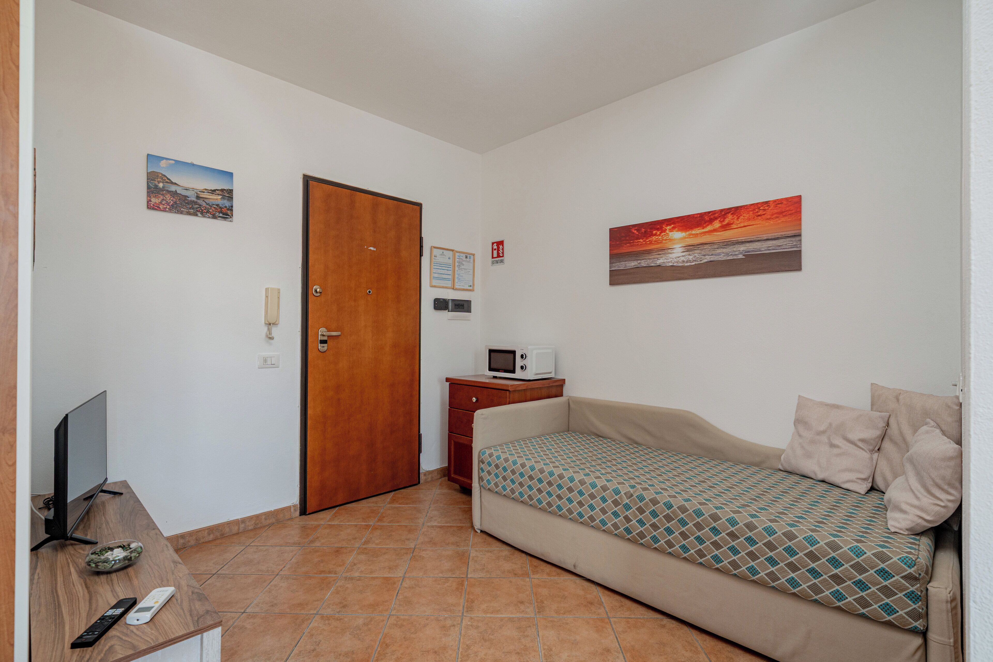 Um quarto pequeno e compacto, com sofá, televisão, porta de madeira e uma pequena mesa com micro-ondas.