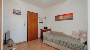 Um quarto pequeno e compacto, com sofá, televisão, porta de madeira e uma pequena mesa com micro-ondas.