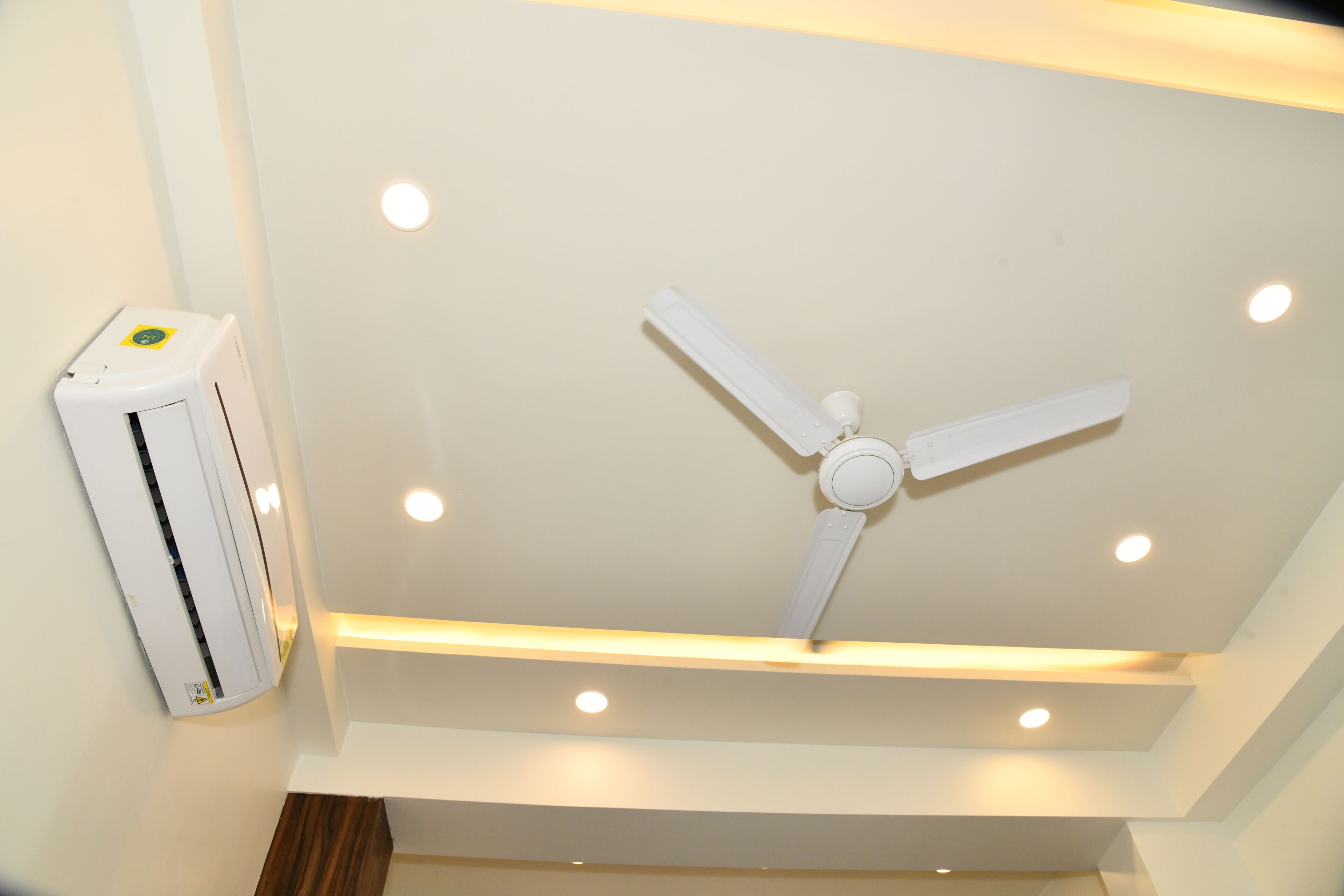 Ventilador de teto com luminária e iluminação embutida em um quarto com paredes brancas e piso de madeira.