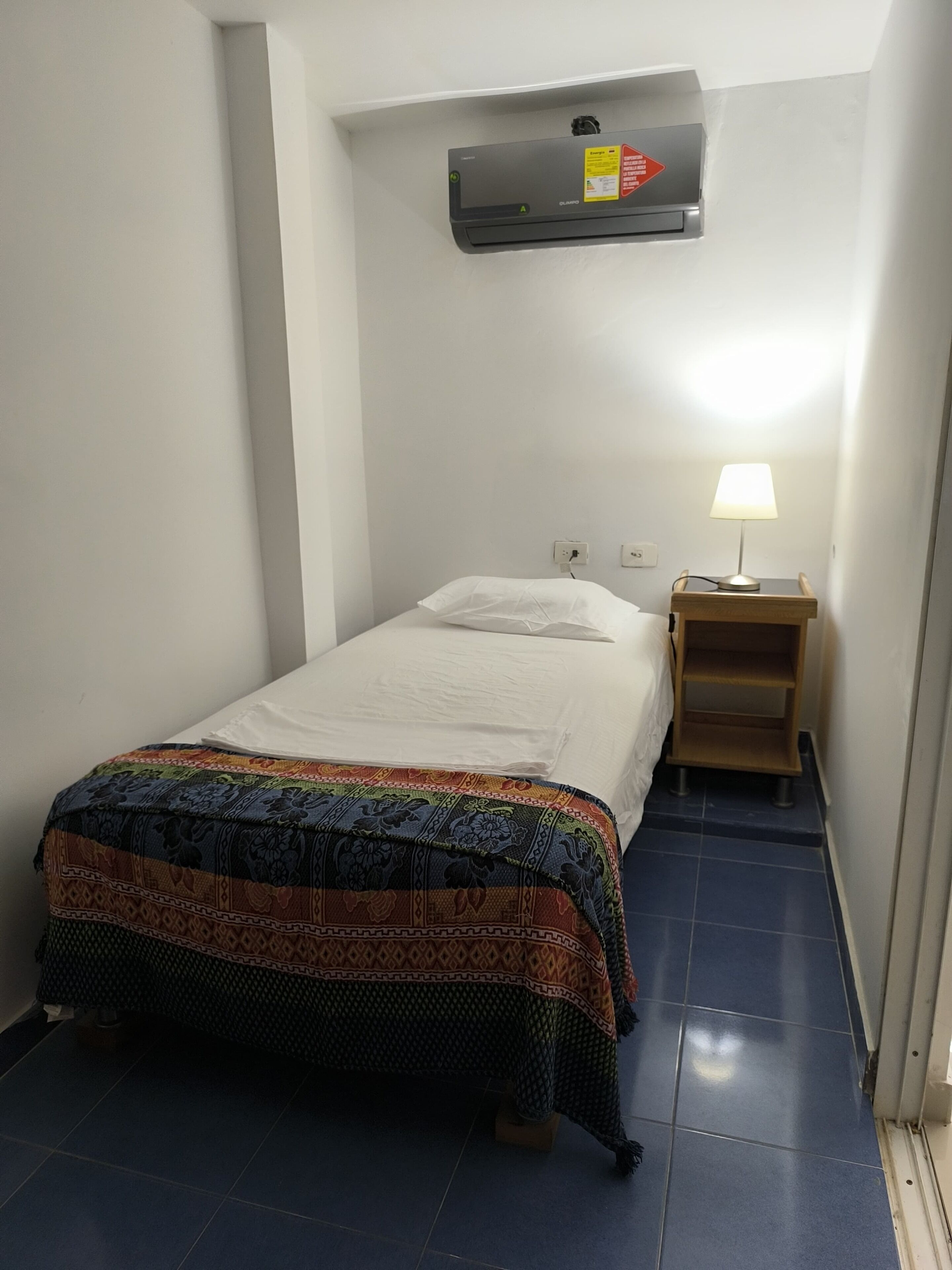 Quarto pequeno com uma cama de solteiro, colcha estampada, luminária de cabeceira e ar-condicionado fixado na parede.