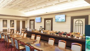 Sala de conferência com uma mesa comprida, cadeiras, serviço de buffet e duas telas na parede.
