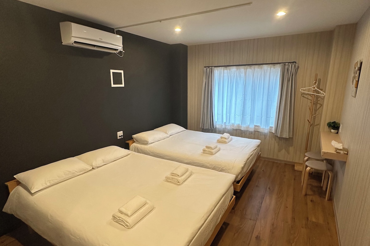 Quarto de hotel com duas camas, janela com cortinas, uma mesinha e um aparelho de ar condicionado.