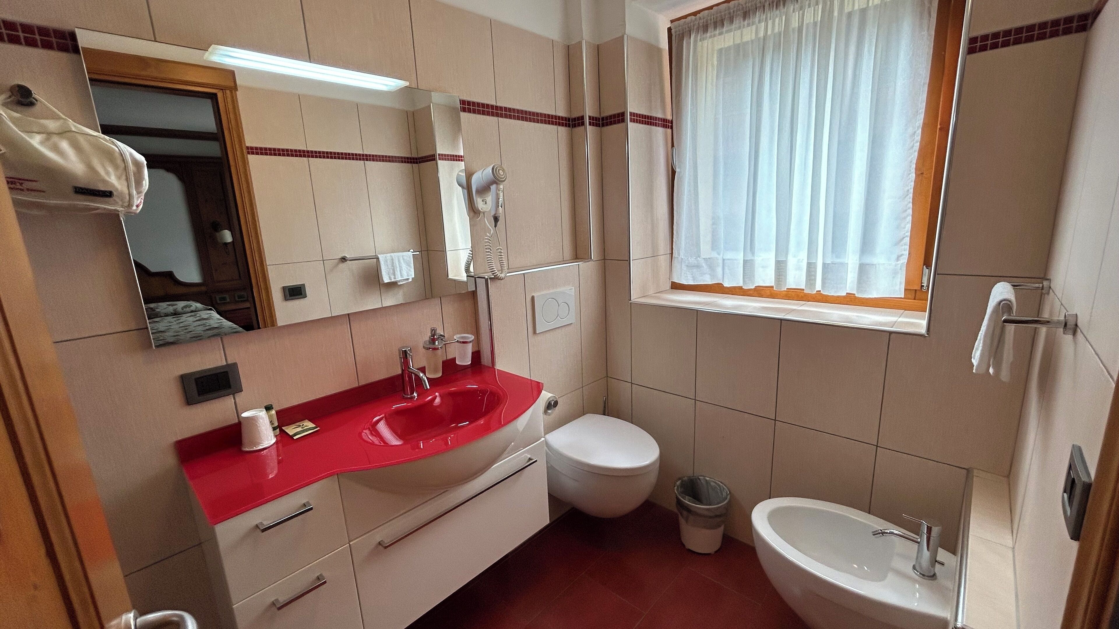 Uma casa de banho com uma pia vermelha, sanita branca e janela com cortinas.